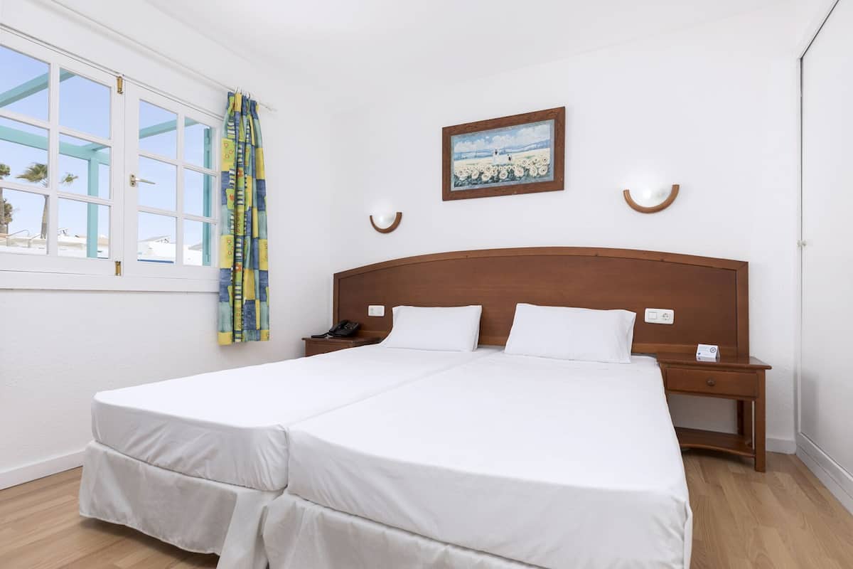 Uma cama de casal com roupa de cama branca, cabeceira de madeira, mesas de cabeceira com luminárias, um quadro na parede e uma janela com cortinas.