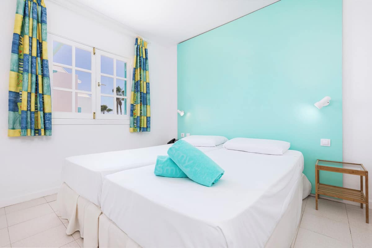 Quarto de hotel com duas camas, uma janela com cortinas, uma parede de destaque azul-esverdeada e uma mesa de cabeceira com abajur.