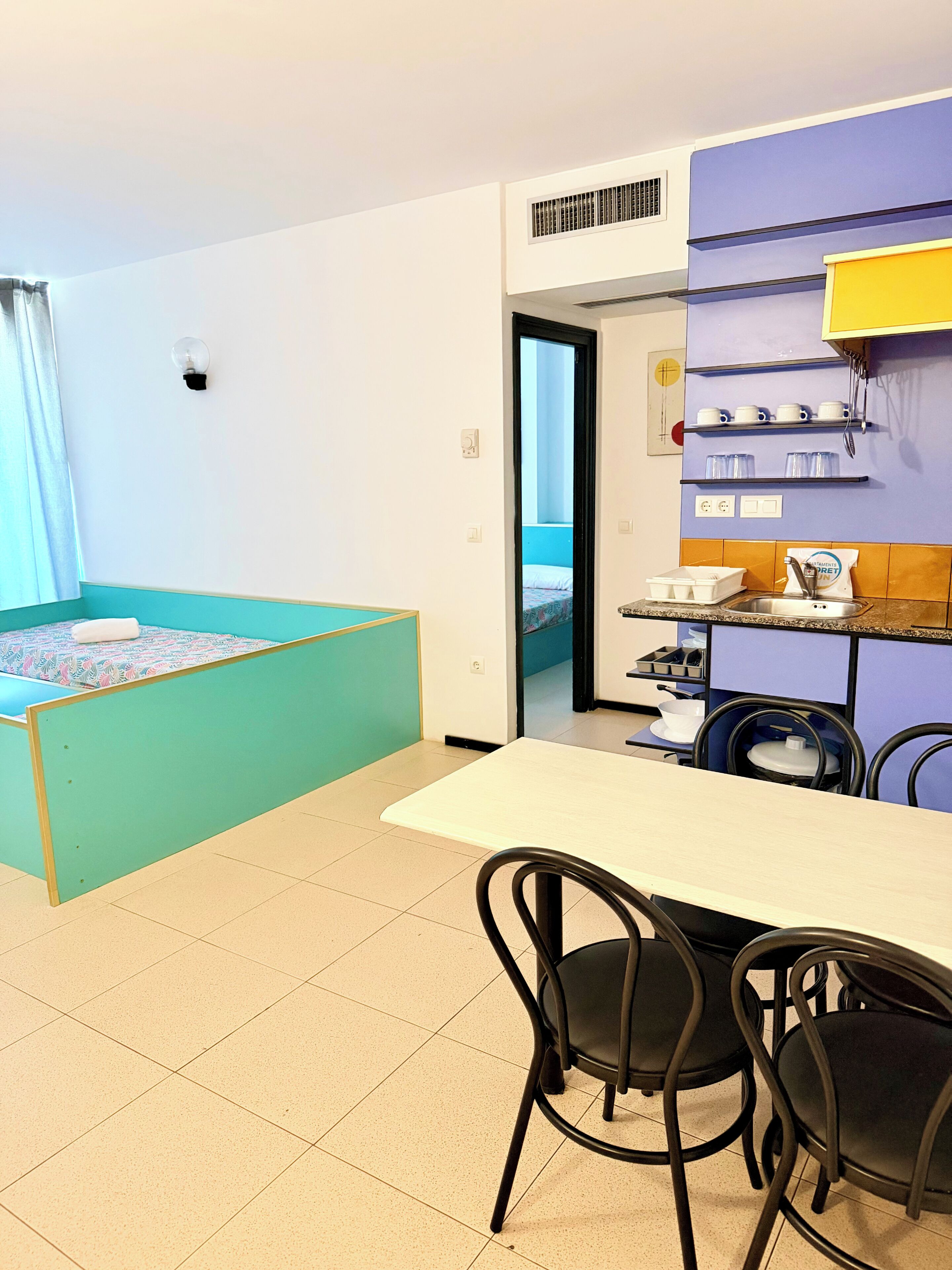 Quarto com cama, mesa e cadeiras, cozinha compacta e banheiro visível por uma porta aberta.