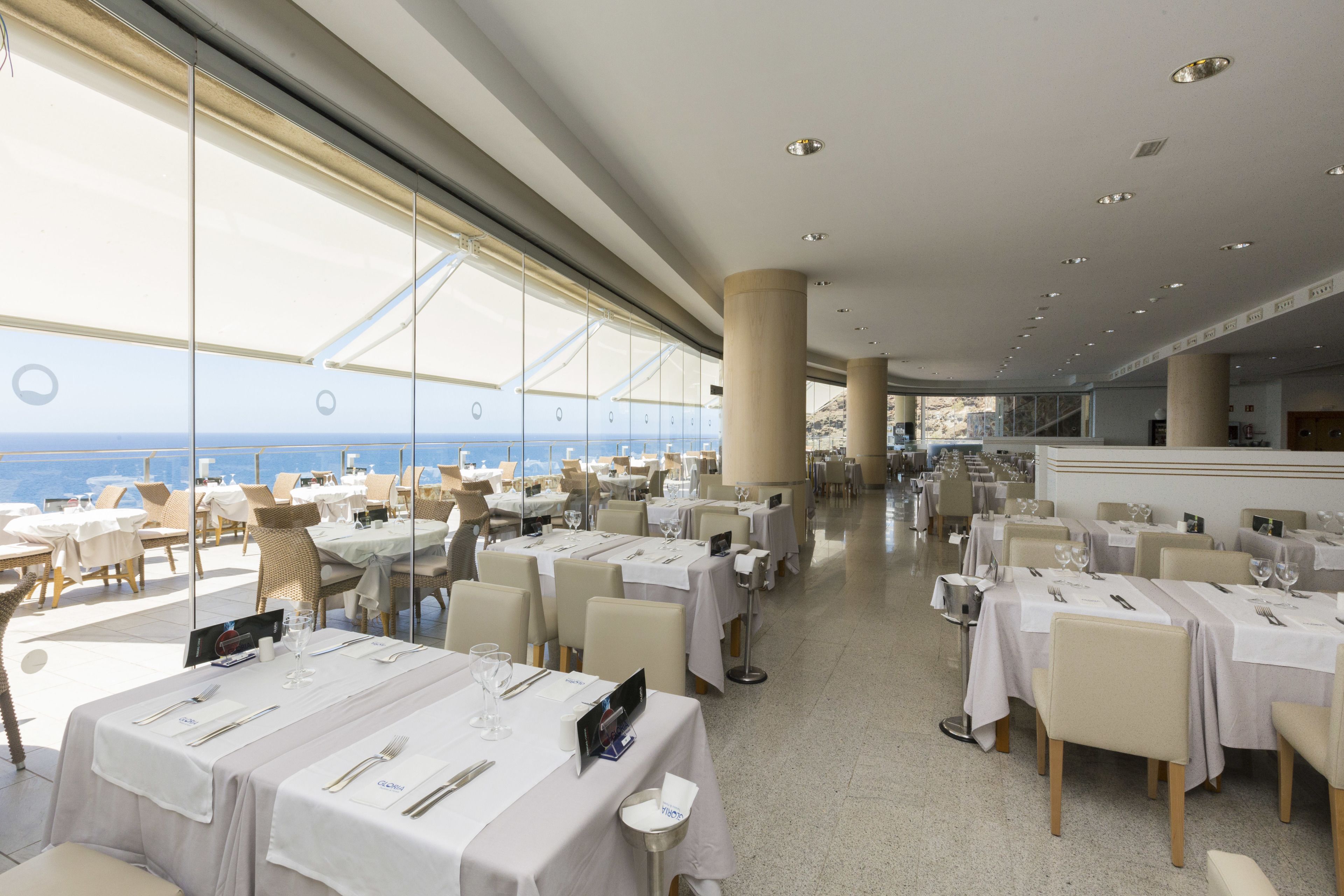 Um restaurante espaçoso com mesas preparadas para refeições, grandes janelas com vista para o mar e um design de interiores moderno.