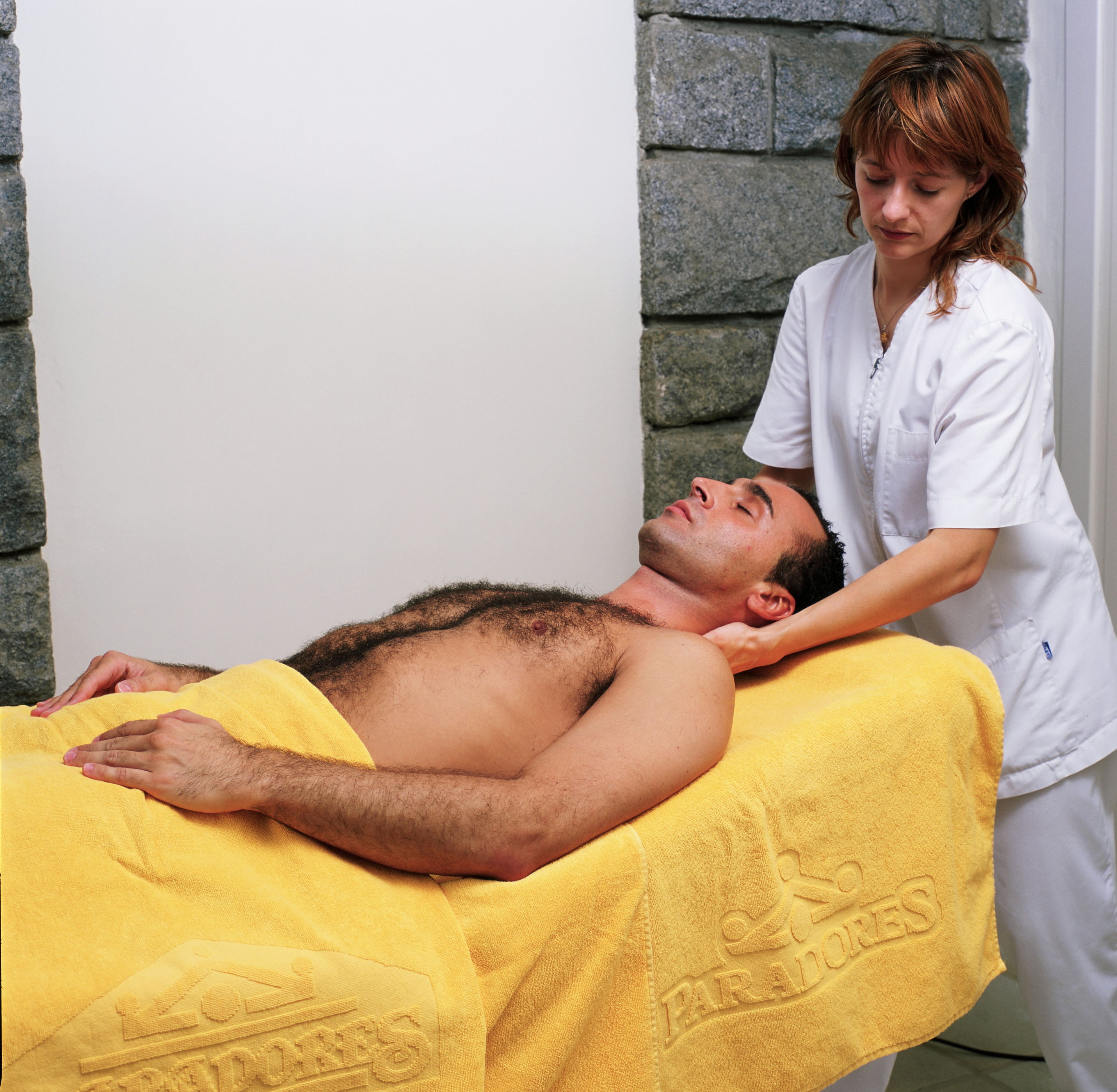 Um homem recebendo massagem em um quarto com paredes de pedra e uma porta branca simples.
