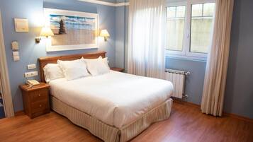 Cama bem arrumada em quarto com cabeceira de madeira, mesa de cabeceira, mesa lateral, quadro na parede, janela com cortinas e radiador.