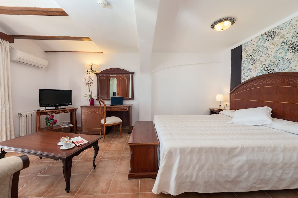Quarto de hotel com uma cama, uma escrivaninha com televisão, uma cadeira, uma mesinha com uma flor e uma mesinha de cabeceira com abajur.