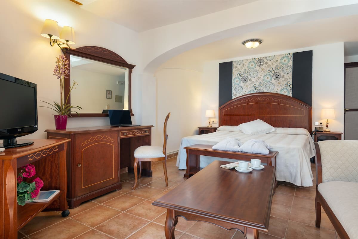 Quarto de hotel com cama de madeira, escrivaninha com televisão, cadeira, mesa pequena e planta em vaso.