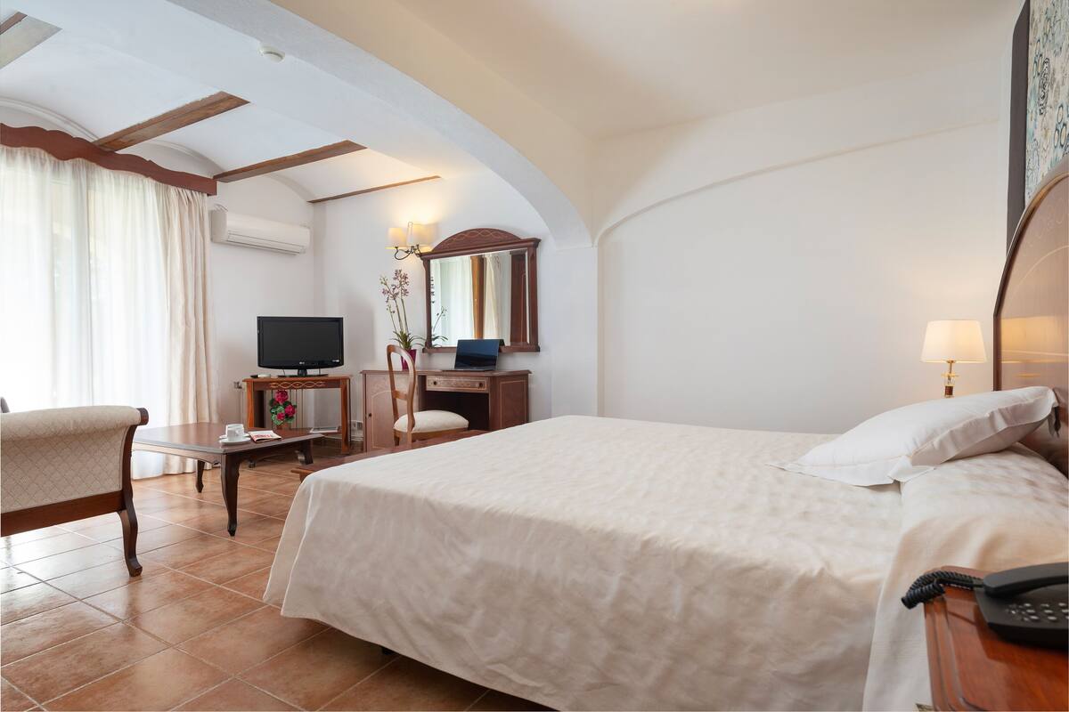 Quarto de hotel com uma cama grande, uma escrivaninha com computador, uma cadeira, uma televisão e um abajur.