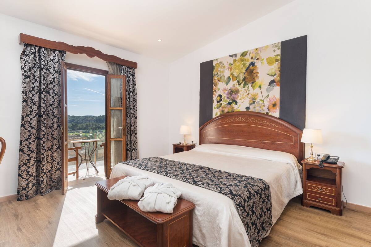 Quarto de hotel com uma cama grande, cabeceira de madeira, mesa de cabeceira com abajur, um banco com toalhas e uma varanda com cortinas.