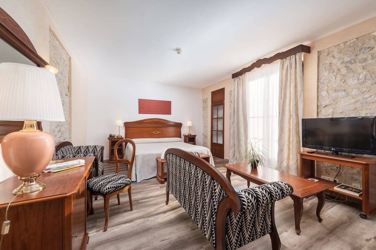 Quarto de hotel com uma cama, uma escrivaninha com luminária, uma televisão e uma cadeira.