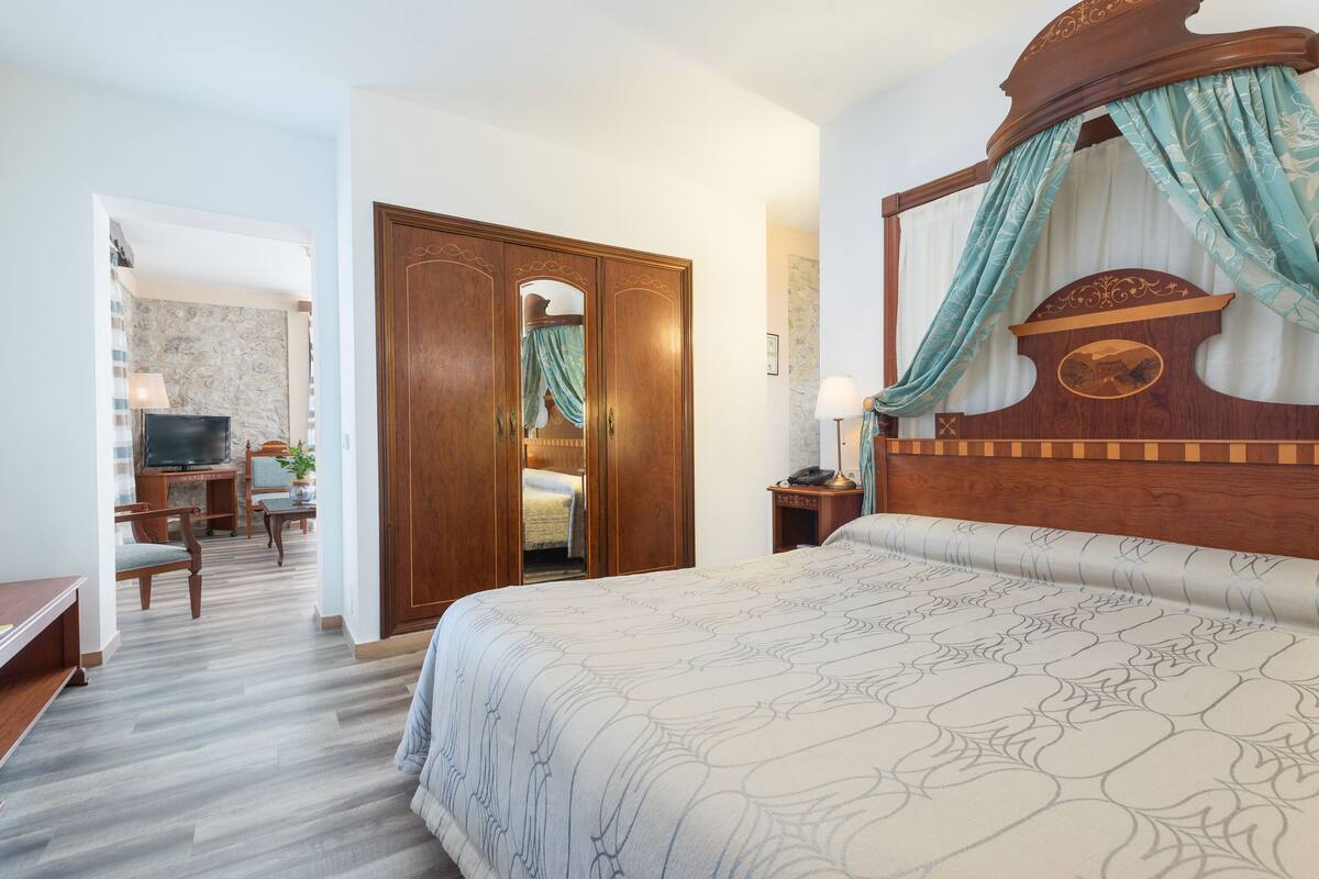 Quarto de hotel com uma cama grande, cabeceira de madeira e um espelho. Há uma escrivaninha com uma cadeira e uma televisão no canto.