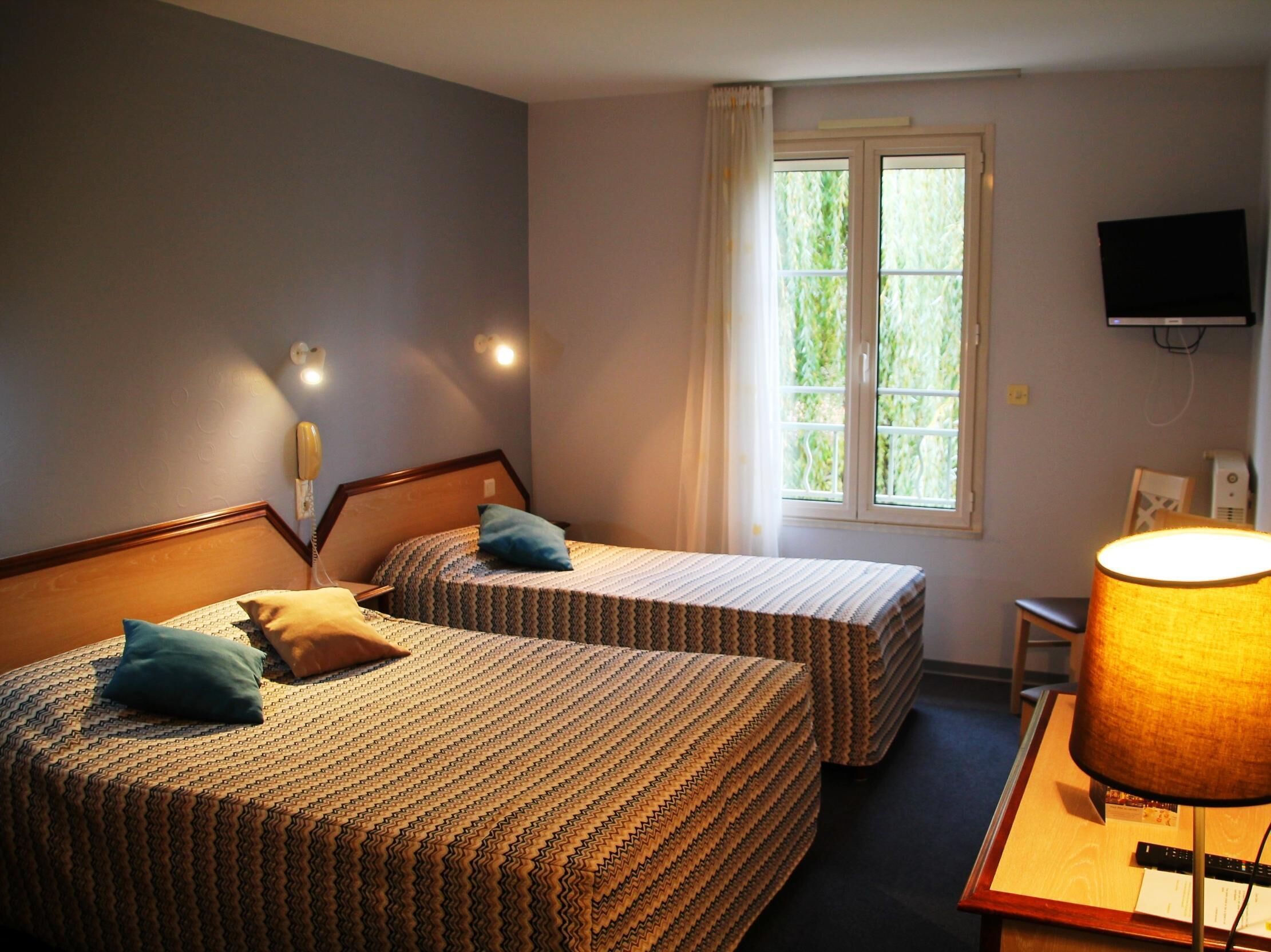 Quarto de hotel com duas camas, uma televisão, uma secretária com candeeiro e uma janela com cortinas.