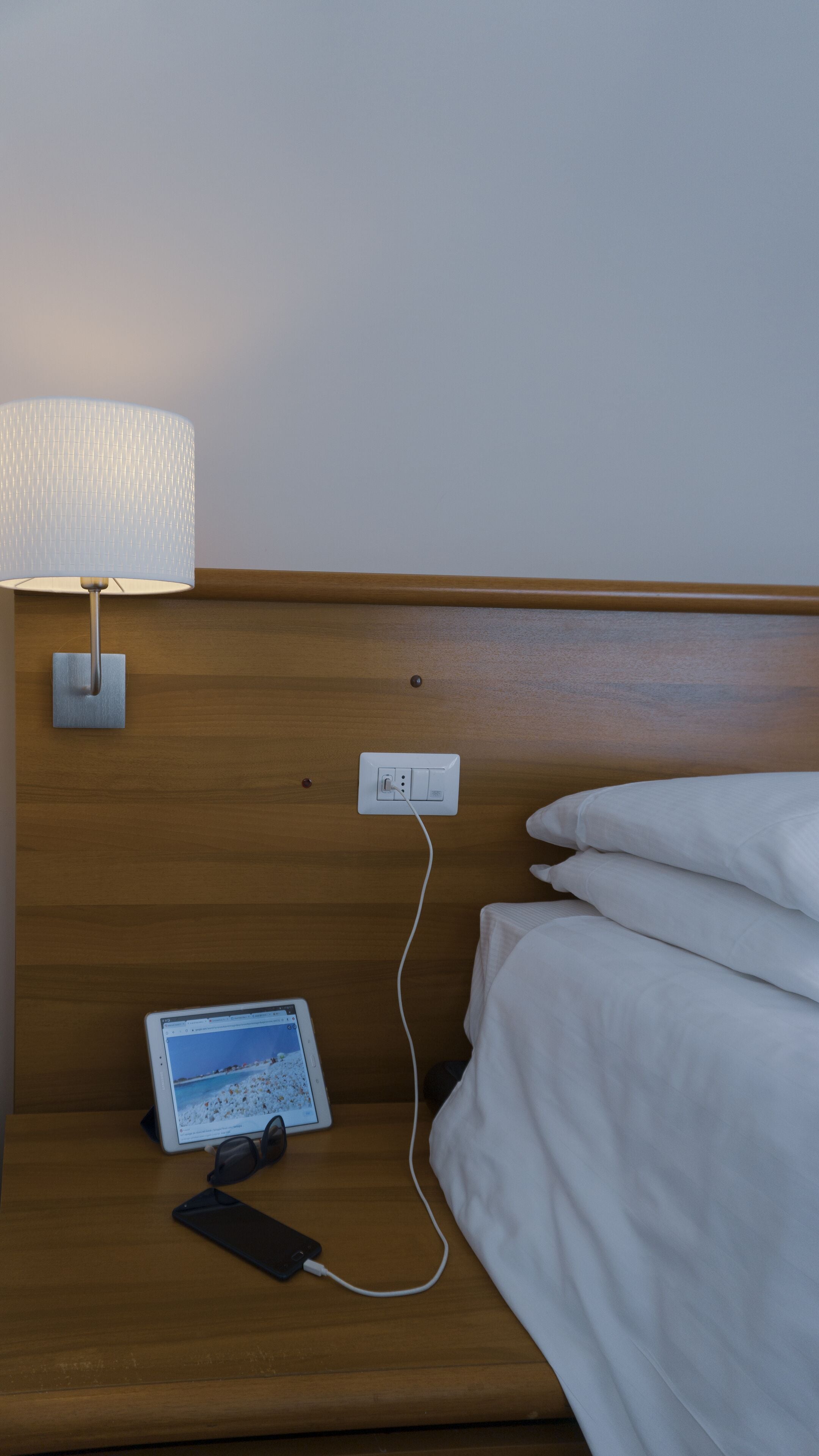 Quarto de hotel com cabeceira de madeira, mesa de cabeceira com abajur, tomada, tablet e carregador de celular.