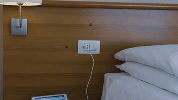 Quarto de hotel com cabeceira de madeira, mesa de cabeceira com abajur, tomada, tablet e carregador de celular.