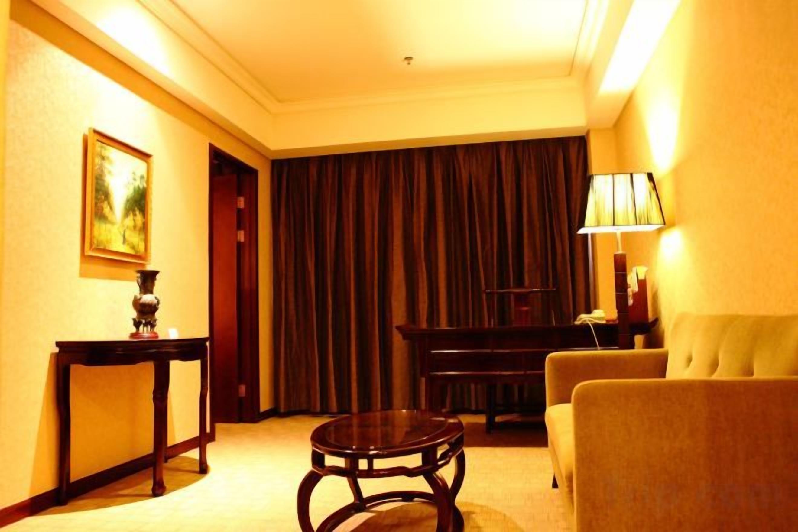 Quarto de hotel com sofá, mesa redonda pequena, luminária e um quadro na parede.