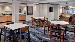 Uma sala de jantar de hotel com mesas e cadeiras redondas, uma cozinha ao fundo e um cartaz grande na parede.