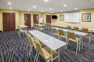Uma sala de conferências com várias mesas e cadeiras, um quadro branco e uma tela de projetor.