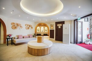 Um lobby moderno com área de estar circular em madeira, um sofá com almofadas, um objeto decorativo na parede e uma porta com a inscrição 'Banho Turco'.