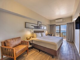 Quarto de hotel com cama, cadeira de madeira, mesa de cabeceira, luminária e uma grande janela com vista.