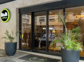 Entrada de hotel com uma grande porta de vidro, plantas em vasos e uma placa com a inscrição 'B&B HOTELS'.