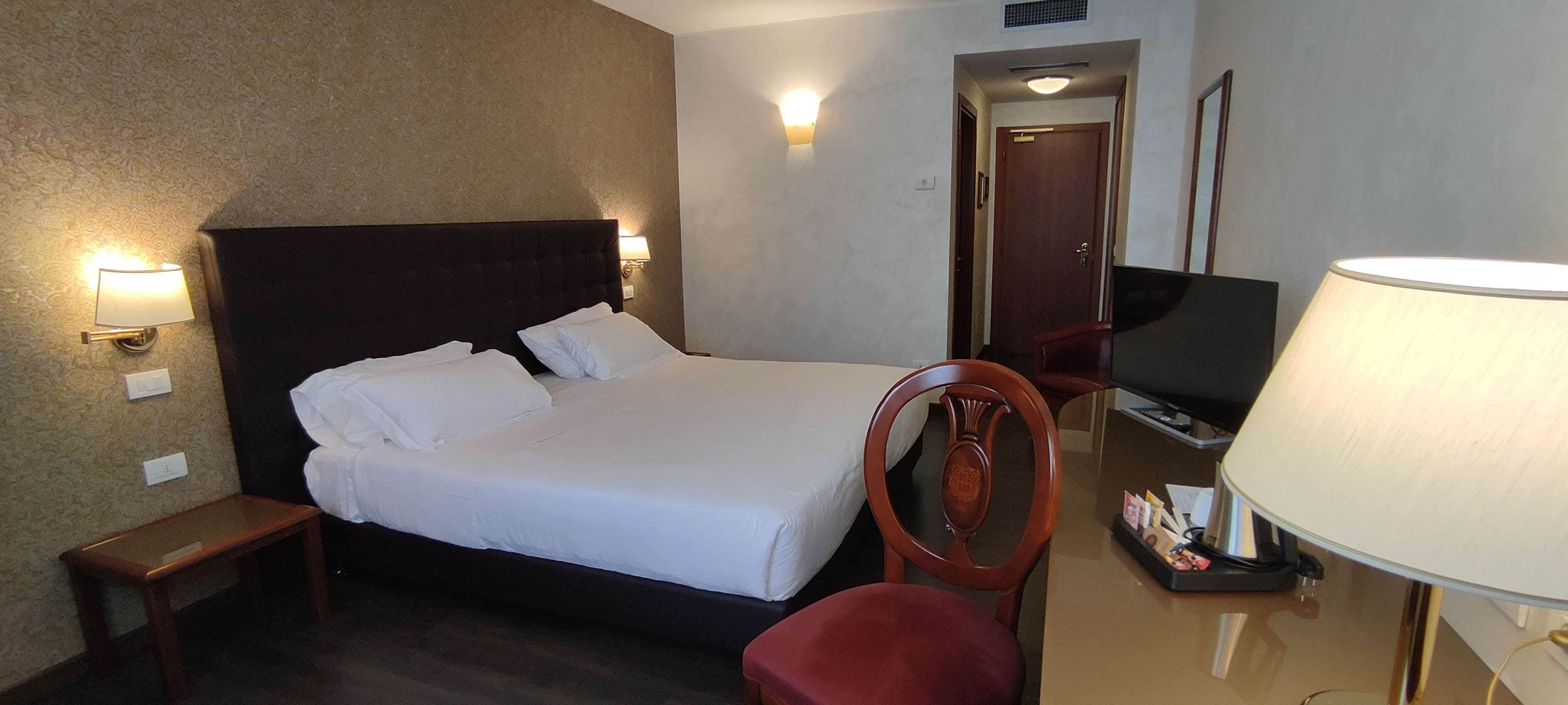 Quarto de hotel com uma cama grande, luminárias de cabeceira, televisão e uma escrivaninha com abajur.