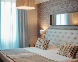 Quarto de hotel com uma cama grande, cabeceira estofada, papel de parede estampado, abajur e janela com cortinas.