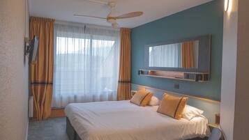 Quarto de hotel com cama, ventoinha no teto, janela grande com cortinas, televisão presa à parede e mesa de cabeceira.