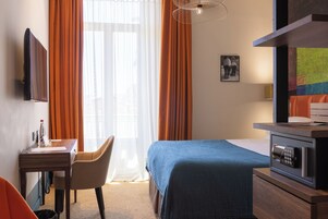 Quarto de hotel com cama, escrivaninha, cadeira, televisão e janela com cortinas.