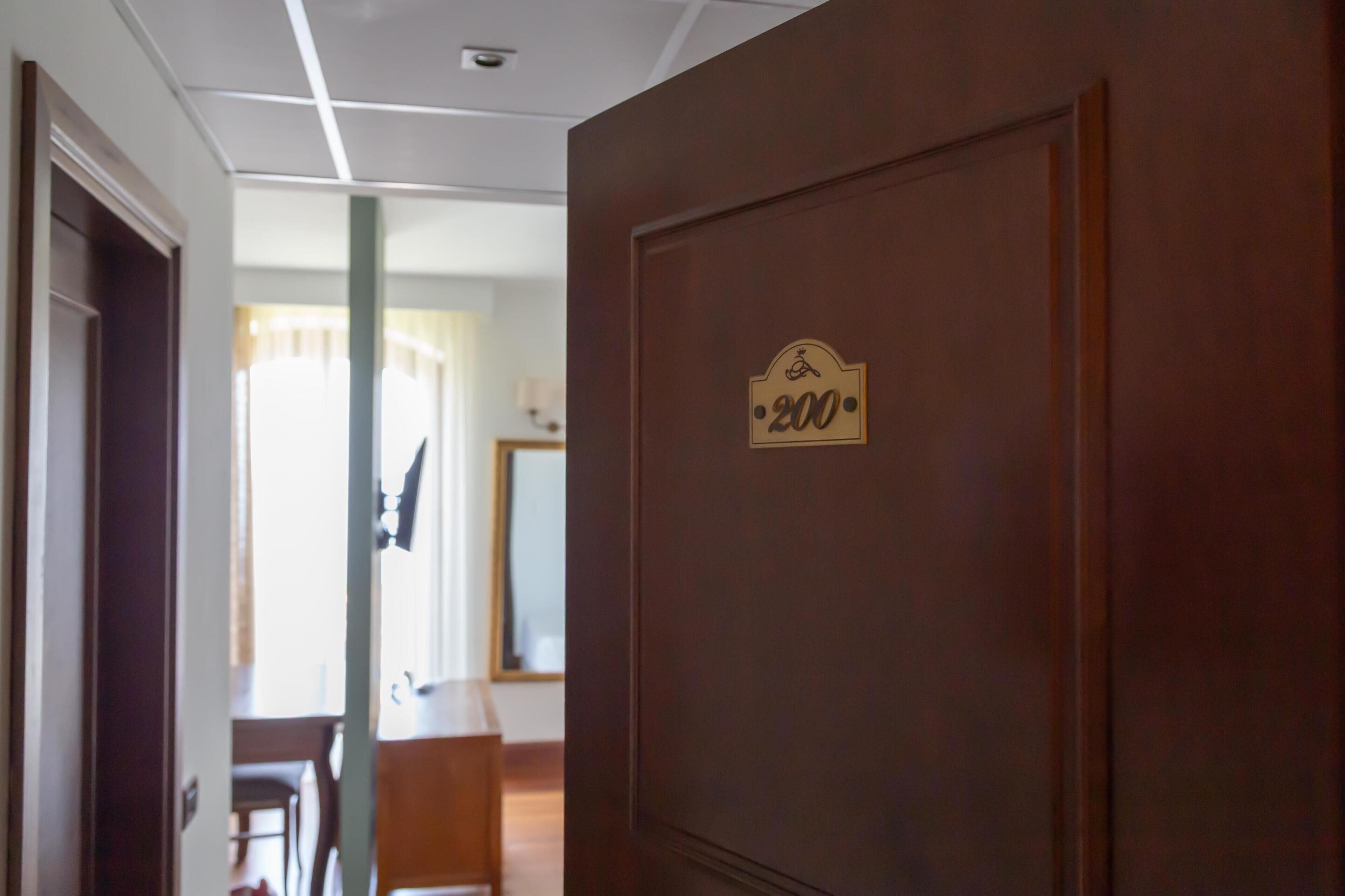 Quarto de hotel com porta de madeira marcada com o número 200, janela visível e uma cadeira ao fundo.