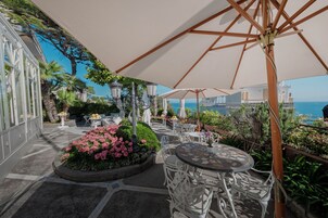 Uma área de terraço ao ar livre com mesas e cadeiras, rodeada por vegetação e vista para o mar.