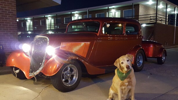 Carro antigo estacionado em frente a um edifício, com um cão sentado ao lado.