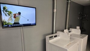 Uma área de lavanderia com uma televisão exibindo uma partida de golfe, uma máquina de lavar e uma secadora.