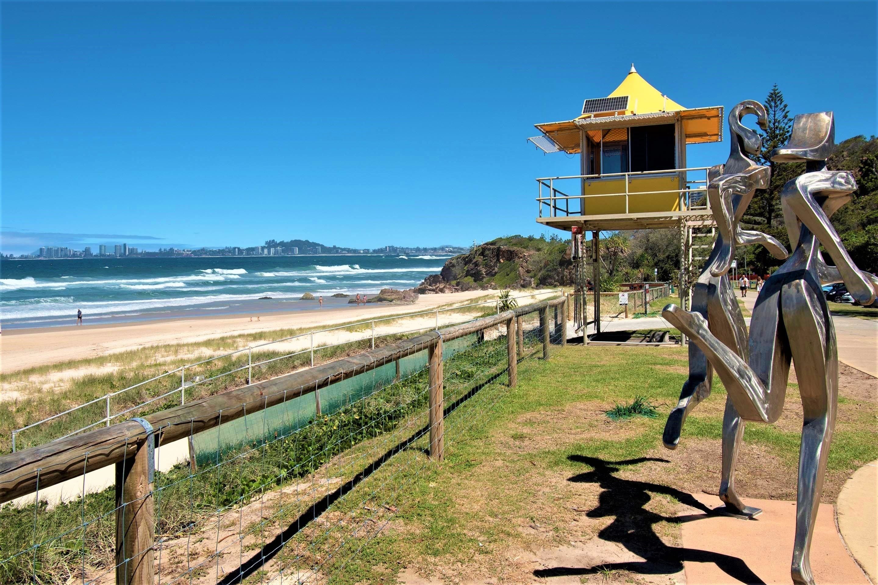 Uma torre de salva-vidas com teto amarelo, com vista para uma praia com ondas e o horizonte de uma cidade ao longe.