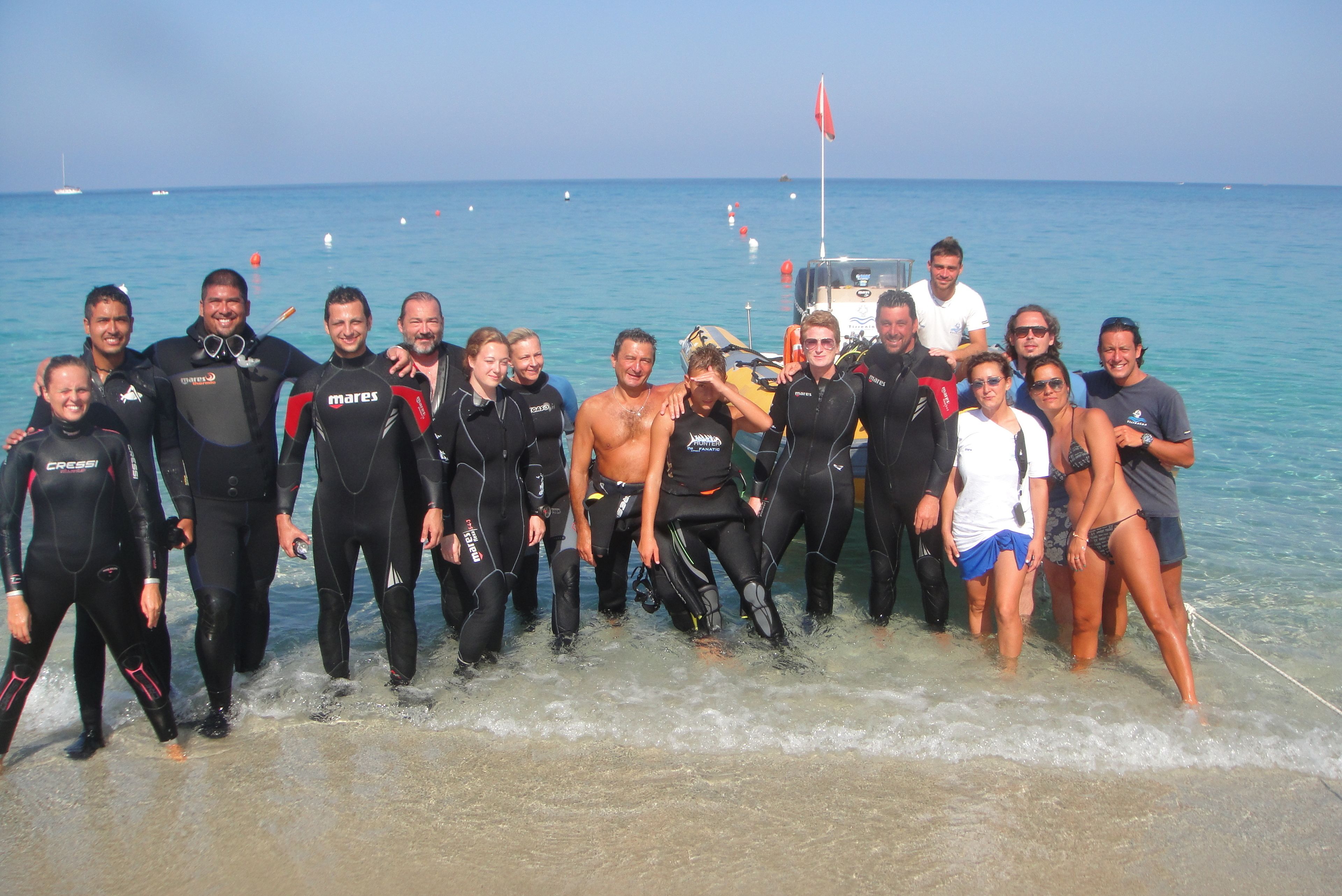 Um grupo de pessoas em fatos de mergulho posando numa praia, com um barco ao fundo.