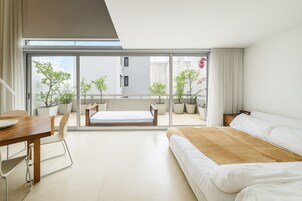 Quarto de hotel moderno com varanda, cama, sofá, mesa de jantar e plantas em vasos.