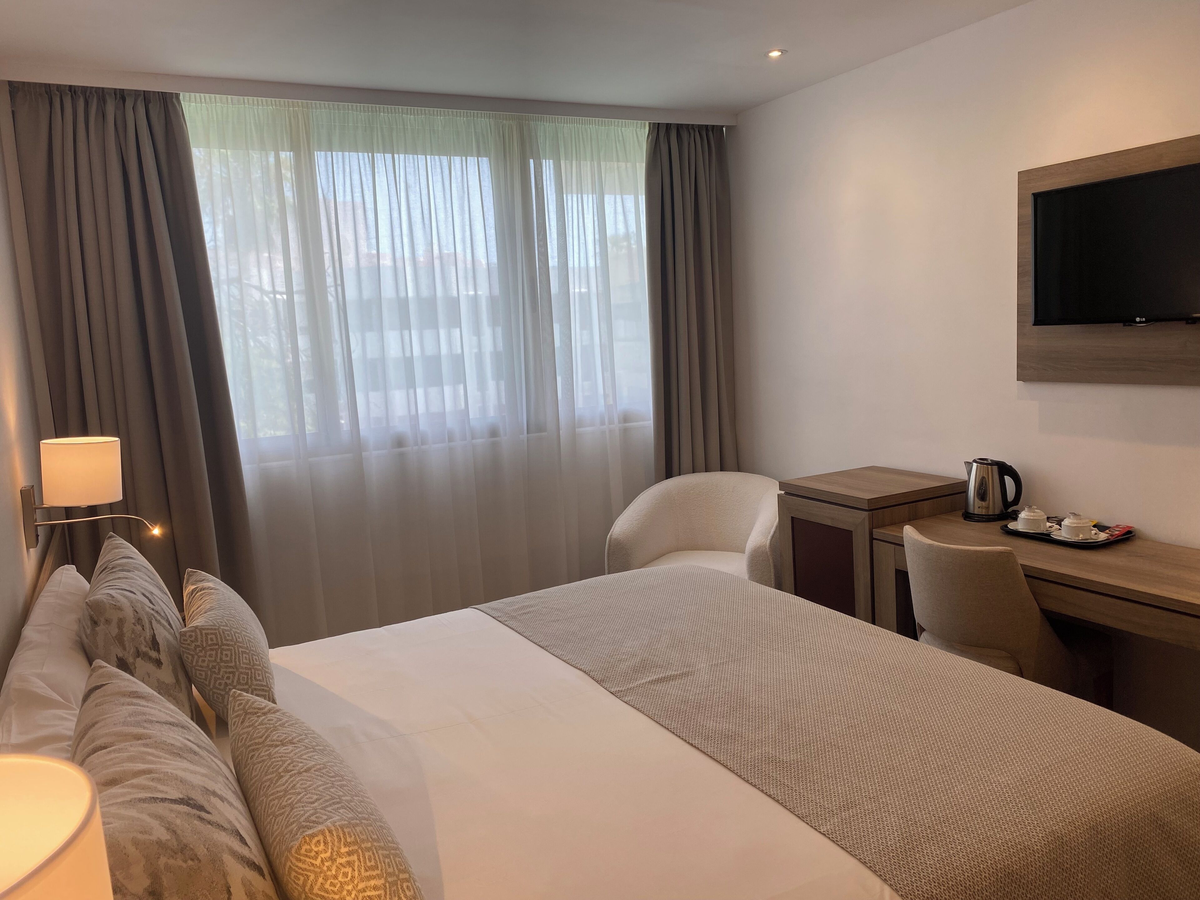 Quarto de hotel com cama, escrivaninha, cadeira, televisão e janela com cortinas.