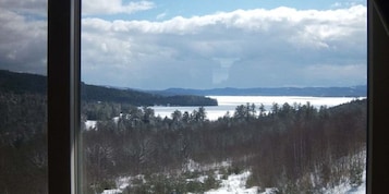 Vista da janela sobre uma paisagem nevada com um lago e colinas arborizadas.