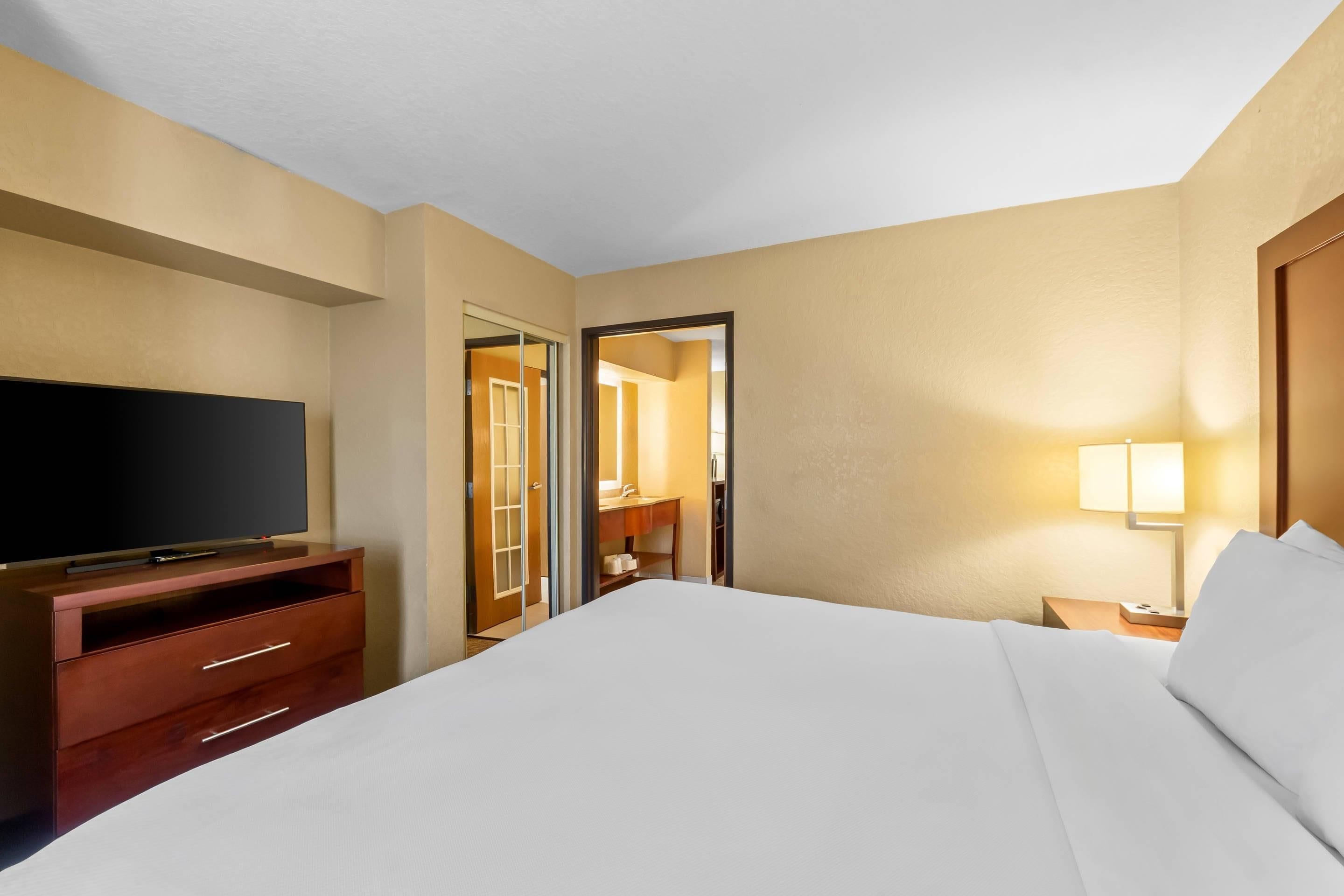 Quarto de hotel com uma cama, uma TV sobre uma cômoda de madeira, uma mesinha de cabeceira com abajur e uma porta que dá para o banheiro.