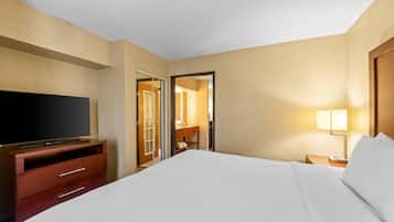 Quarto de hotel com uma cama, uma TV sobre uma cômoda de madeira, uma mesinha de cabeceira com abajur e uma porta que dá para o banheiro.