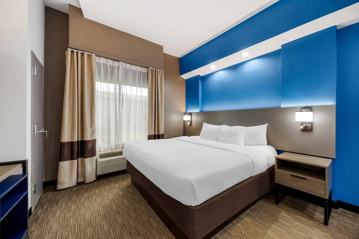 Quarto de hotel com uma cama grande, mesa de cabeceira, luminária na parede e janela com cortinas.