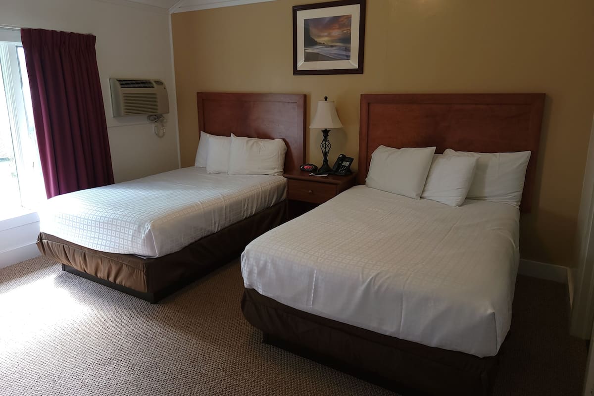 Duas camas de casal com cabeceiras de madeira, uma mesa de cabeceira com abajur e telefone, um quadro na parede e uma janela com cortinas.