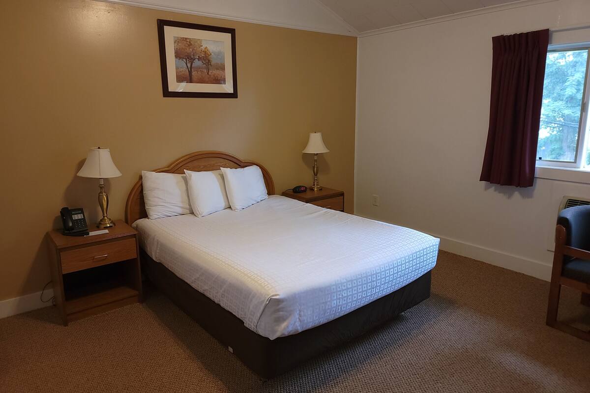 Quarto de hotel com uma cama, duas mesas de cabeceira com luminárias, um relógio, um quadro na parede e uma janela com cortinas.