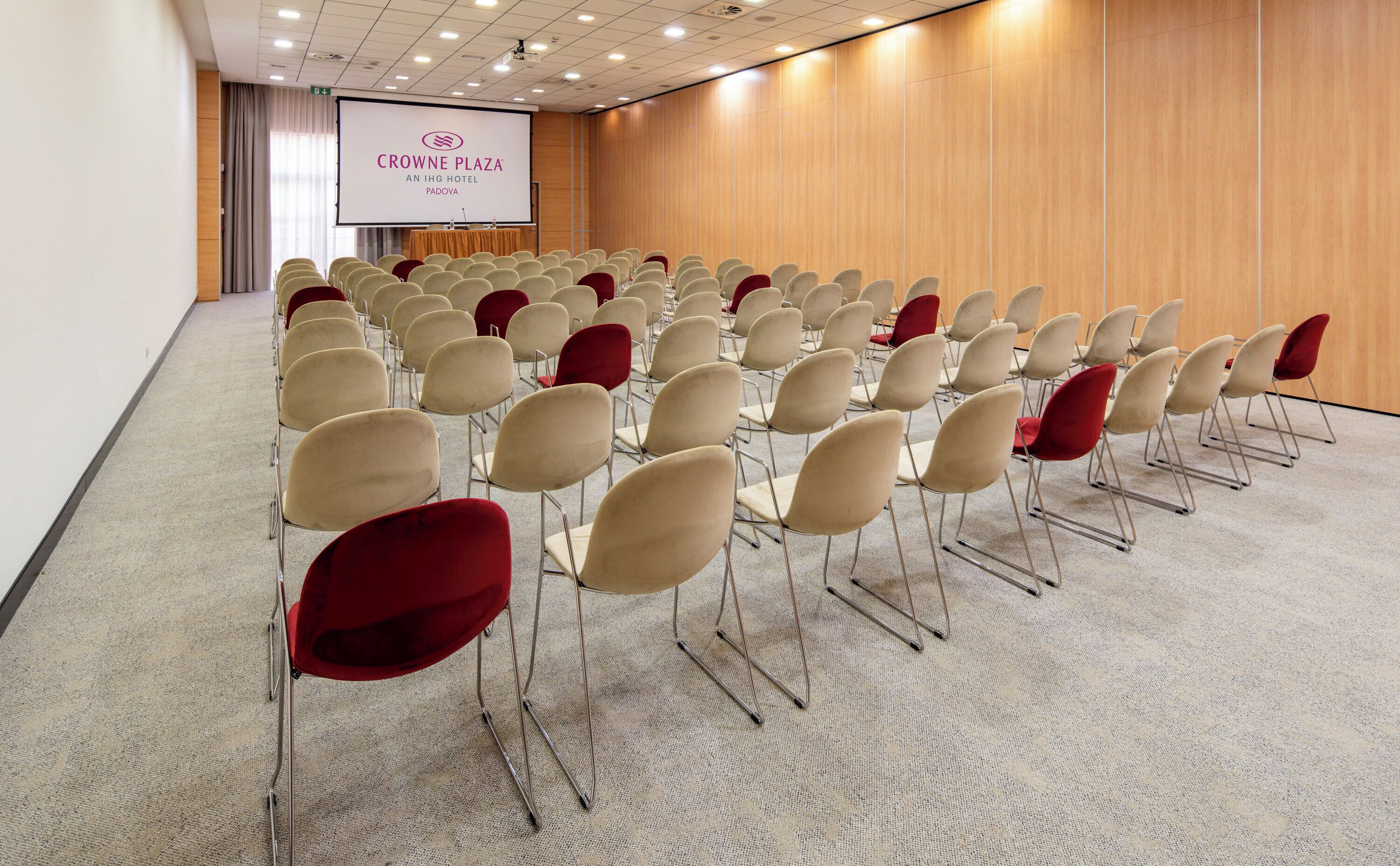 Uma sala de conferências com fileiras de cadeiras bege e vermelhas, um pódio e uma tela de projeção exibindo o logotipo Crowne Plaza.