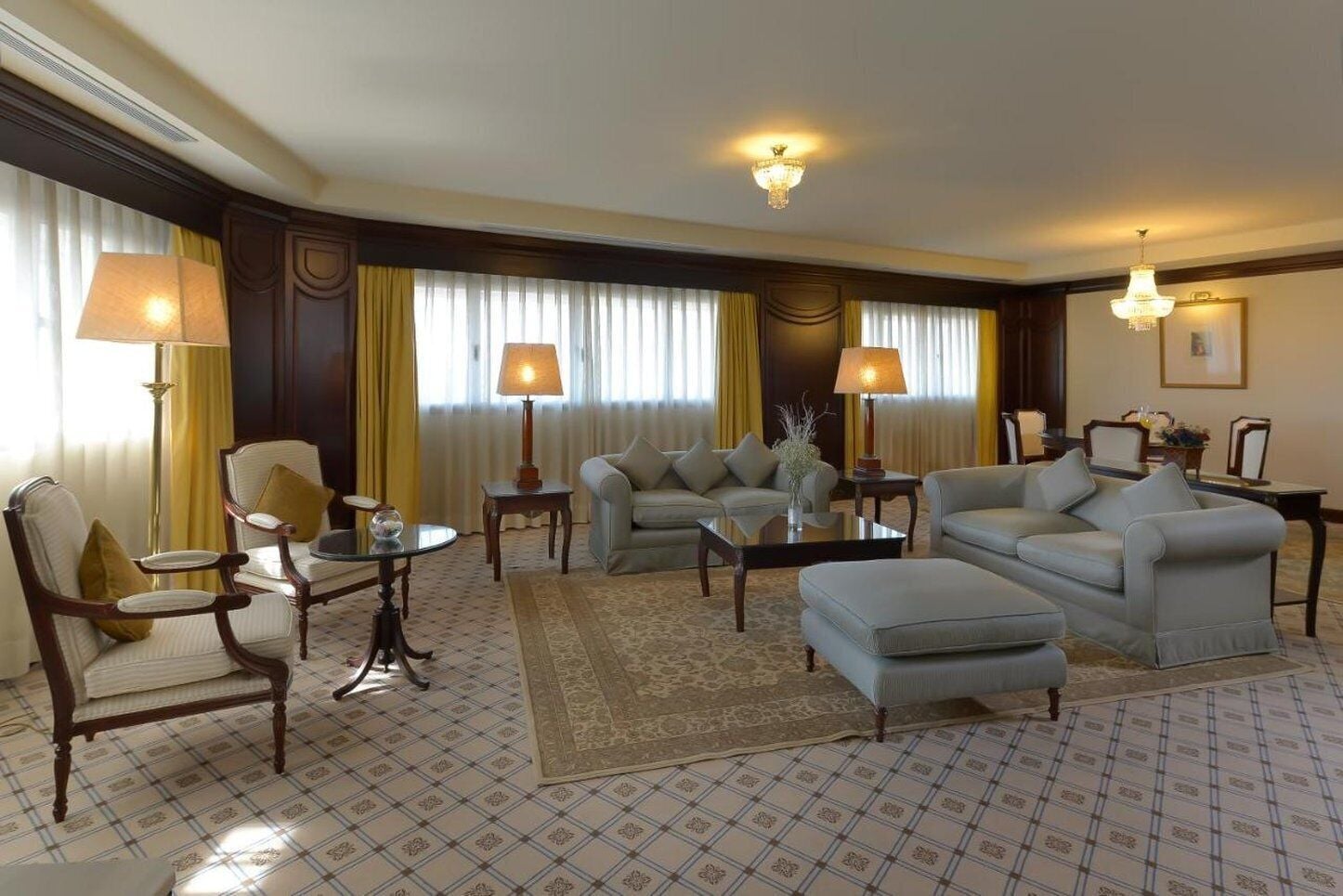 Uma sala de estar ampla com um sofá grande, poltronas, uma mesa de centro e um tapete. Há duas luminárias em mesinhas laterais, e o cômodo apresenta um design clássico com cortinas e um lustre.