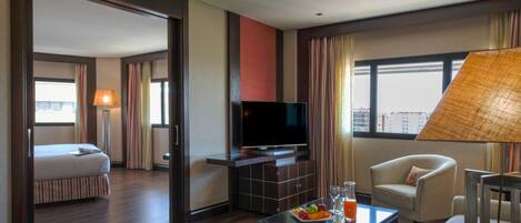 Quarto de hotel com cama, televisão, mesa de jantar com fruta e bebidas, duas poltronas e um abajur.