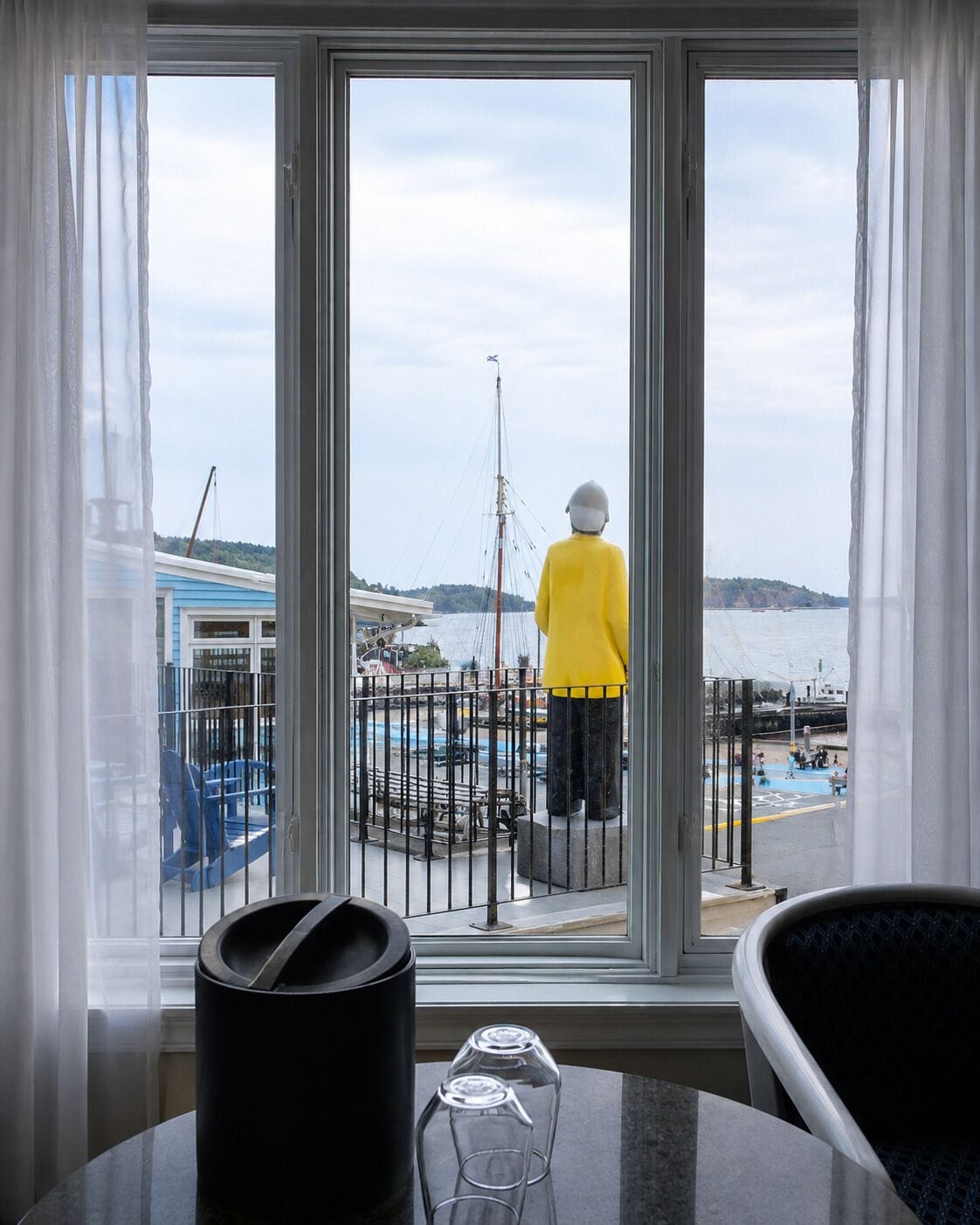 Quarto com uma grande janela com vista para um porto, uma estátua e uma mesa com um objeto preto e três copos.
