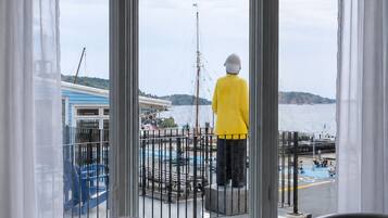 Quarto com uma grande janela com vista para um porto, uma estátua e uma mesa com um objeto preto e três copos.