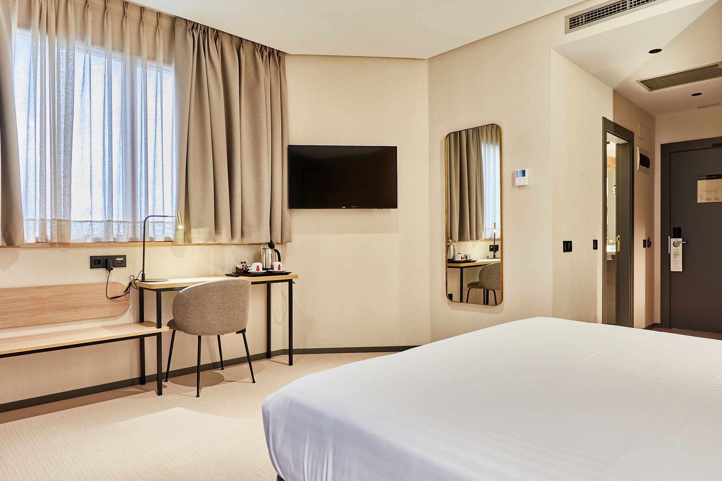 Quarto de hotel com cama, escrivaninha, cadeira, televisão e espelho.
