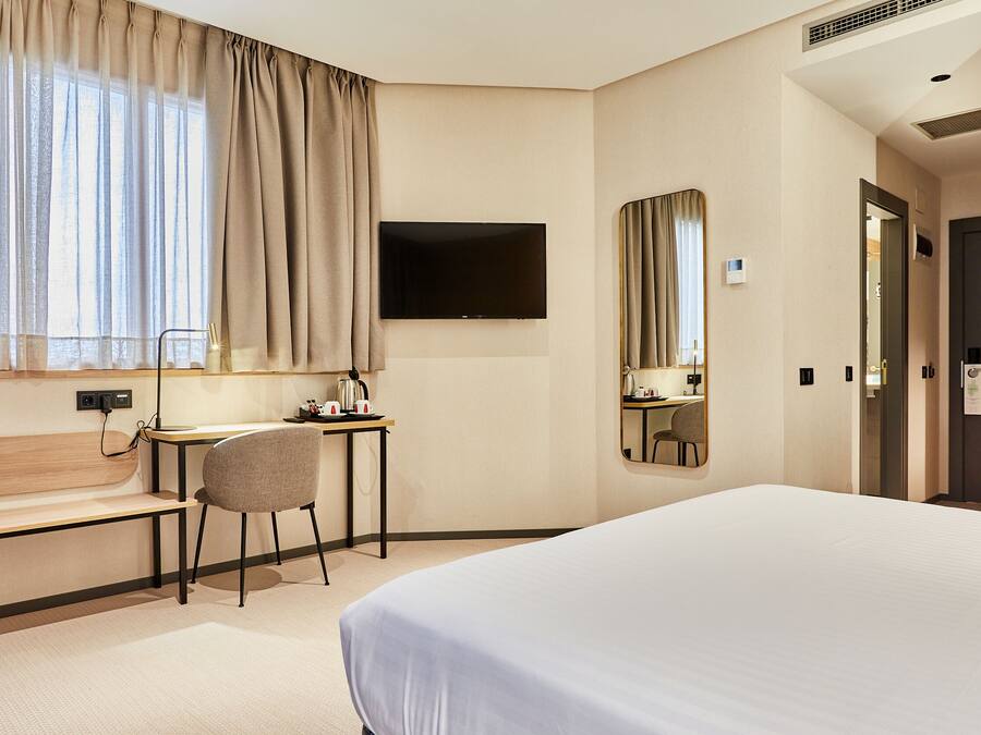 Quarto de hotel com cama, escrivaninha, cadeira, televisão e espelho.