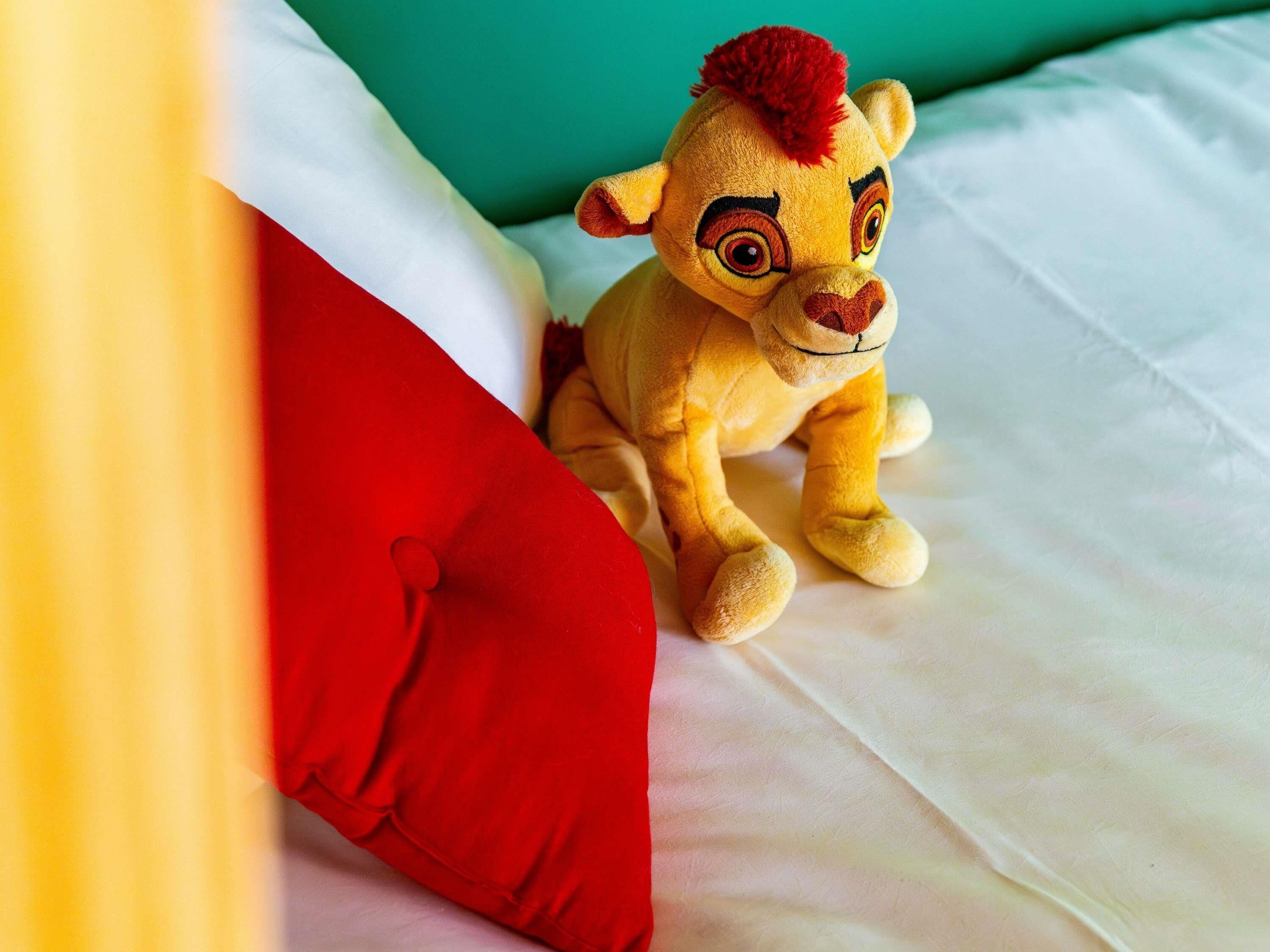 Um brinquedo de pelúcia numa cama com um travesseiro branco e um travesseiro vermelho.