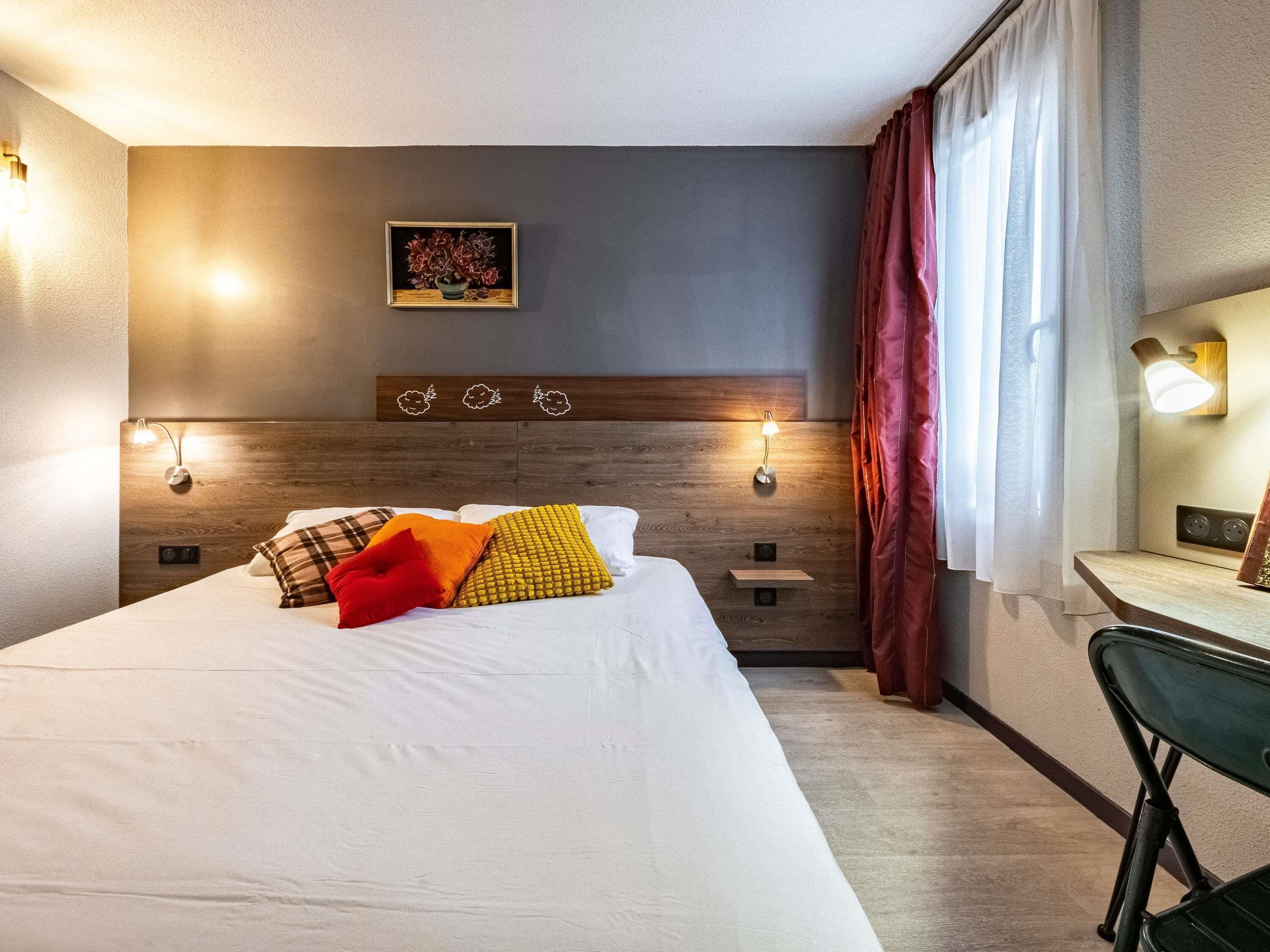 Quarto de hotel com cama, abajures, uma escrivaninha e um quadro na parede.