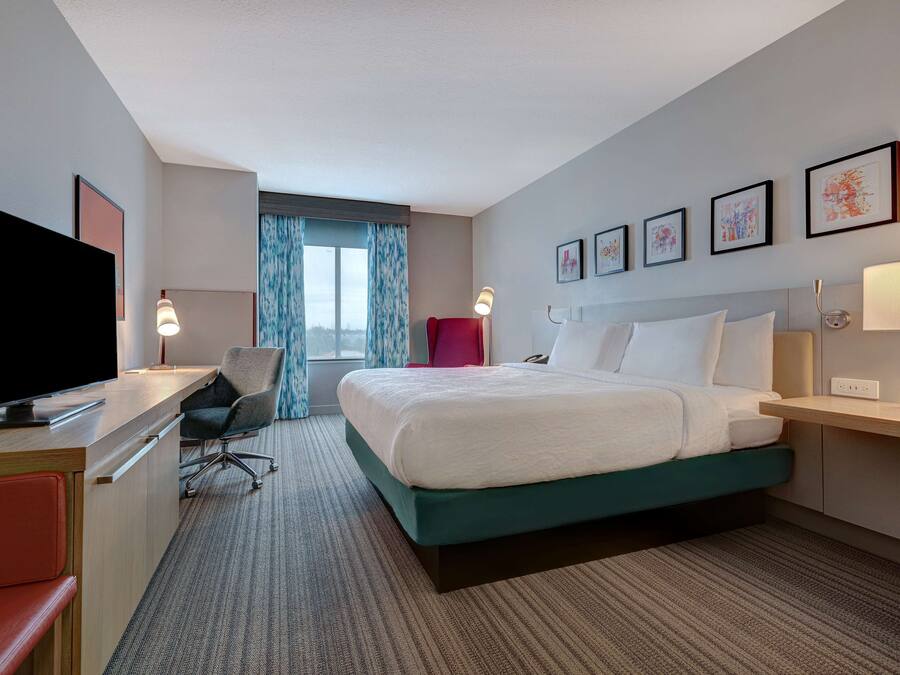 Quarto de hotel com cama, escrivaninha, cadeira, televisão e obras de arte nas paredes.