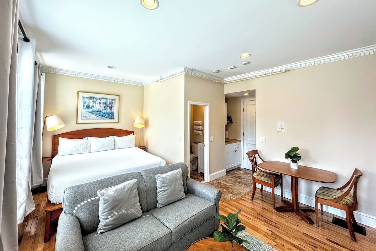 Quarto de hotel com cama, sofá cinzento, mesa de jantar com cadeiras e uma pequena planta.